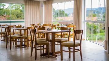 Uma sala de jantar com mesas e cadeiras de madeira, janelas amplas com cortinas, e vista para áreas verdes e colinas.