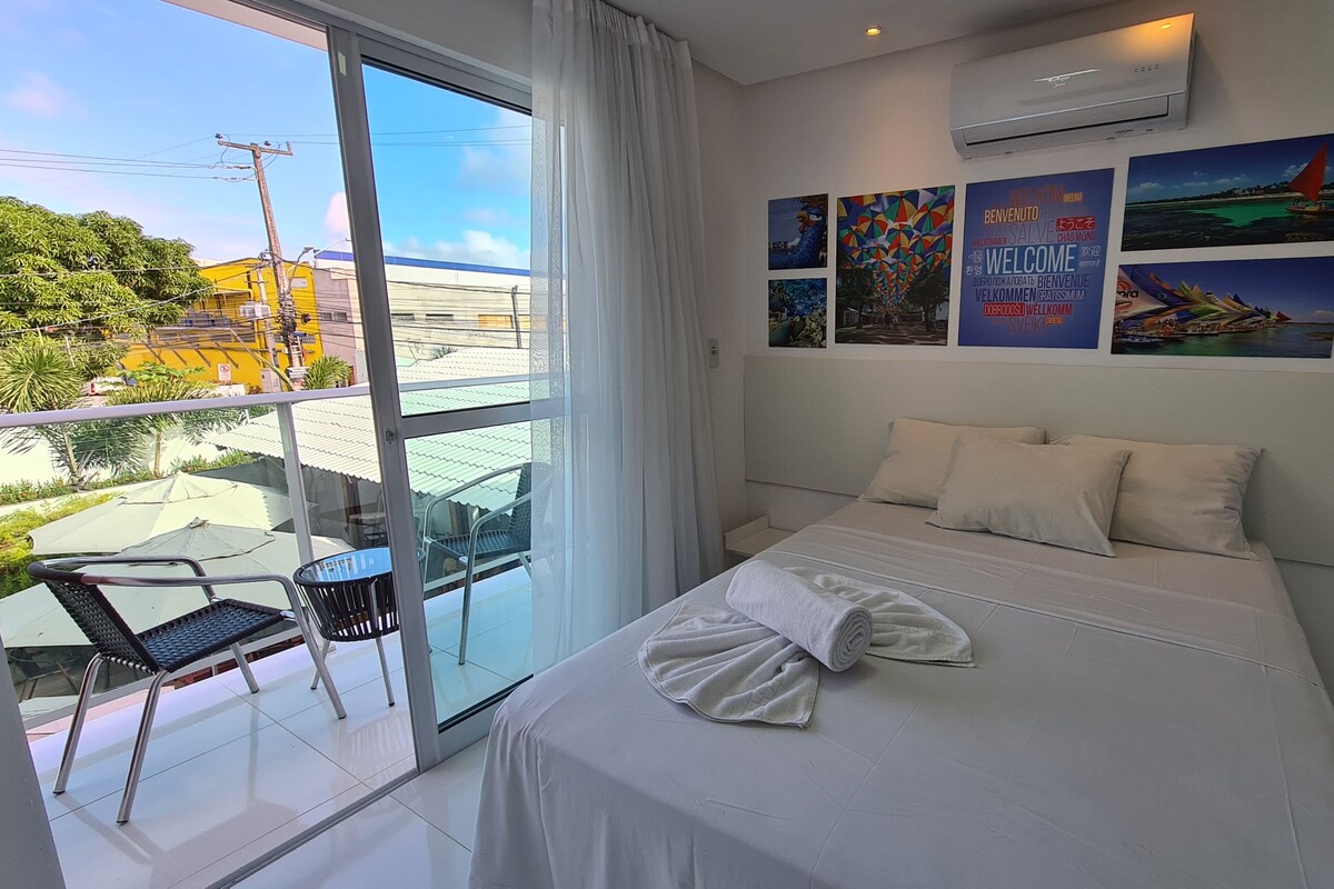 Quarto de hotel com uma cama grande, uma varanda com uma cadeira e vista para a cidade.