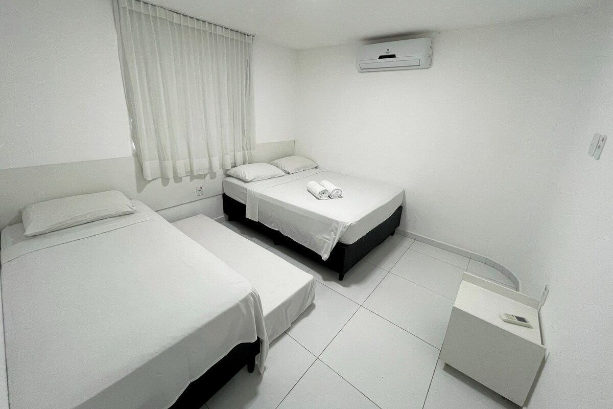 Quarto pequeno e limpo, com duas camas de solteiro, janela com cortinas, ar condicionado fixado na parede e uma pequena mesa de cabeceira.