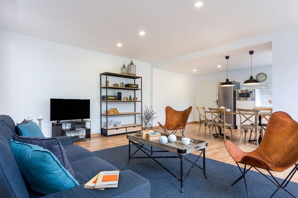 Uma sala de estar moderna com um sofá, mesa de centro e televisão. Há uma área de jantar com cadeiras e uma mesa.