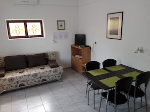 Uma sala de estar pequena com um sofá, uma TV, um armário de madeira e uma mesa de jantar com cadeiras.