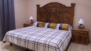 Uma cama bem arrumada com colcha xadrez, duas mesas de cabeceira com luminárias e uma cabeceira de madeira com detalhes florais entalhados.