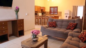 Uma sala de estar com sofá, mesa de centro e TV. Cozinha ao fundo, com armários de madeira e janela.