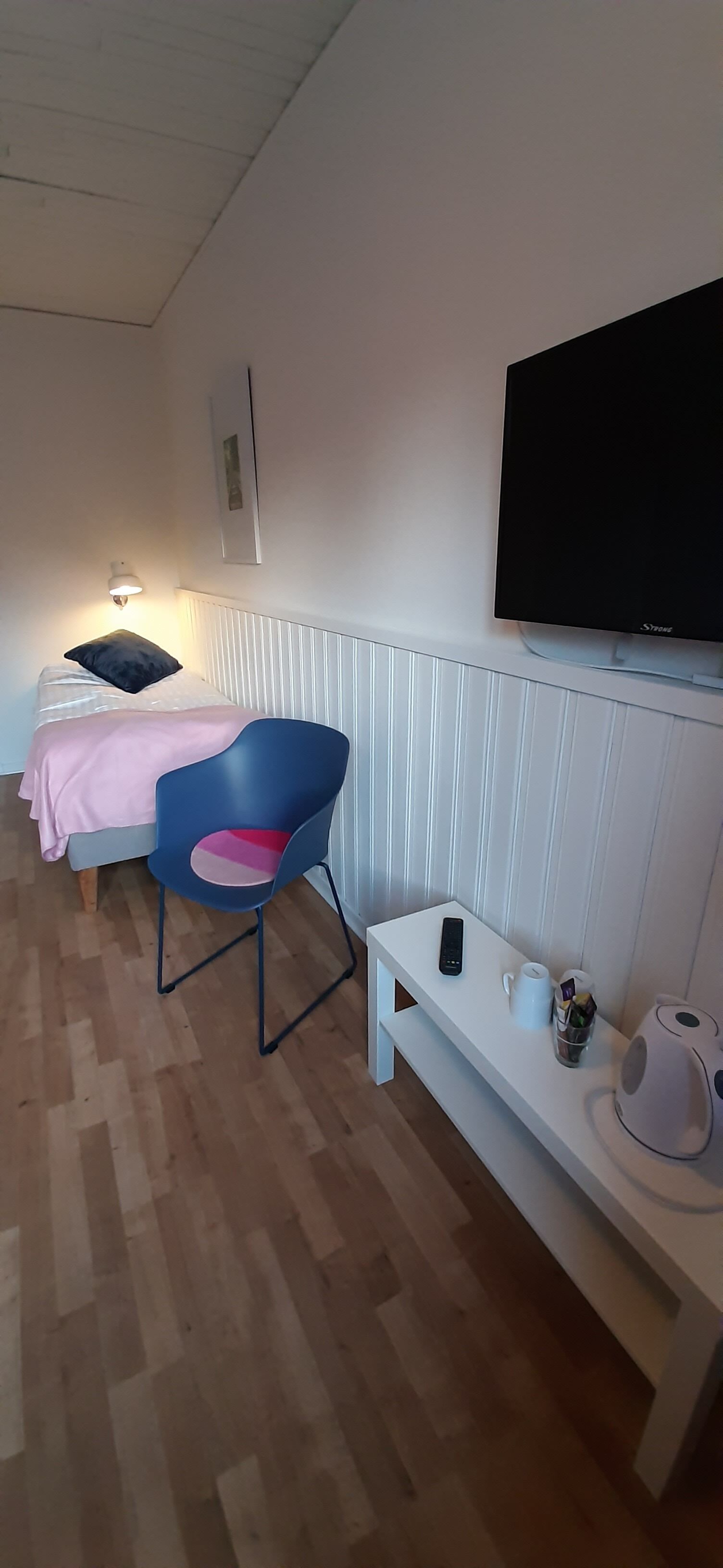 Quarto com cama, cadeira, televisão, mesa de cabeceira com controle remoto e chaleira.