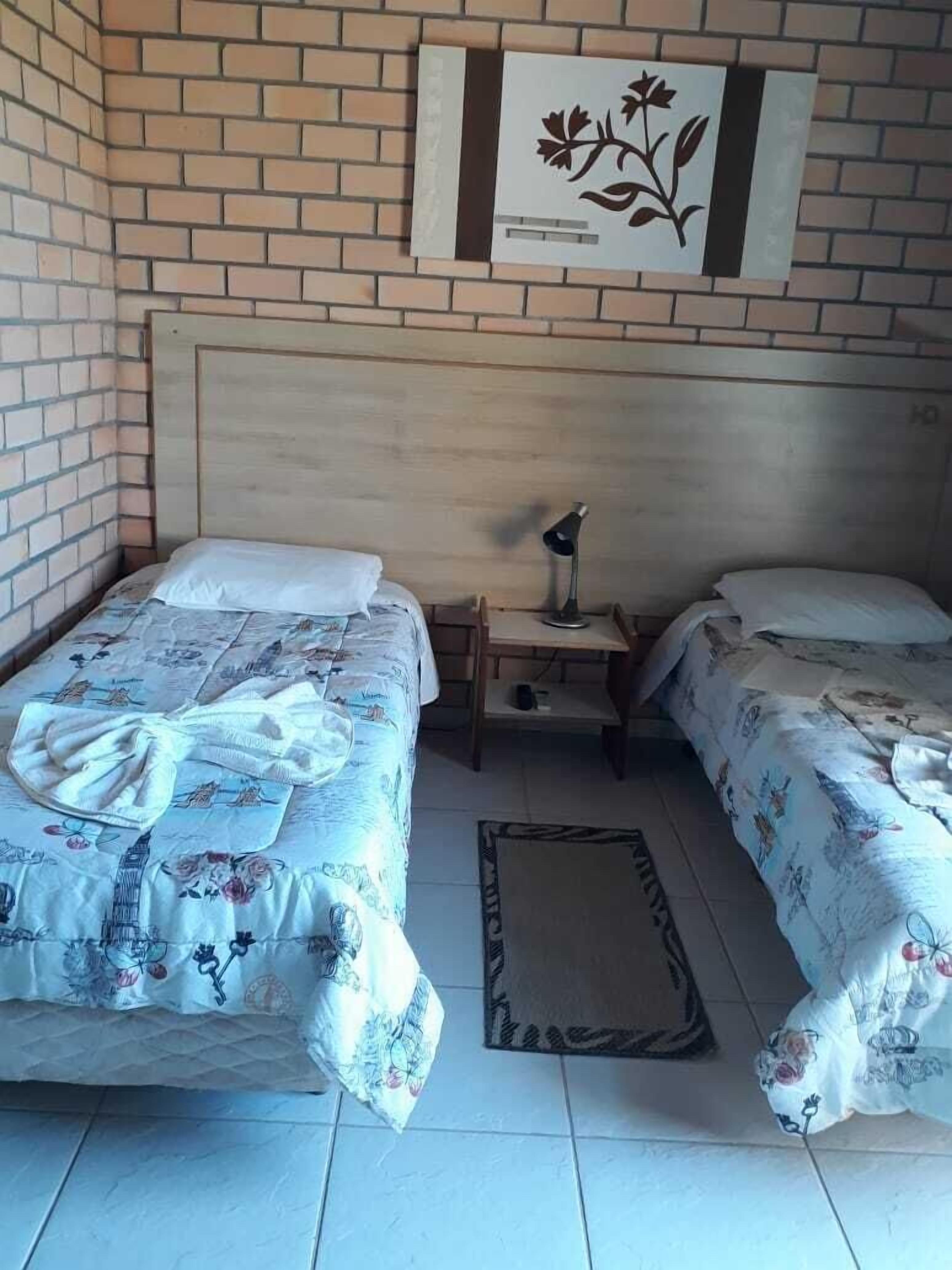 Quarto com duas camas, uma mesa de cabeceira, uma luminária e um objeto decorativo na parede.