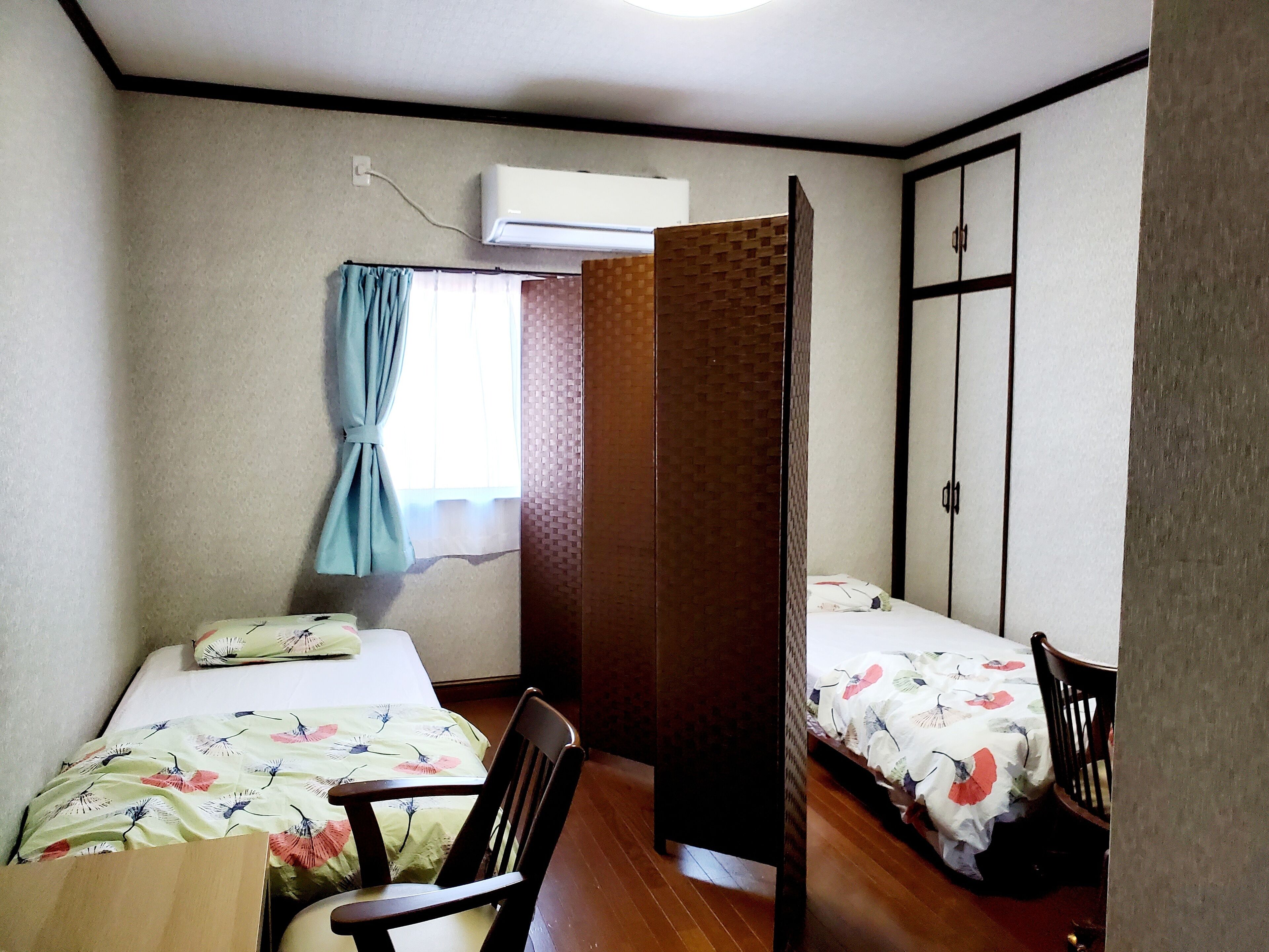 Quarto de hotel pequeno e individual, com duas camas, uma cadeira de madeira, um guarda-roupa e um ar condicionado.