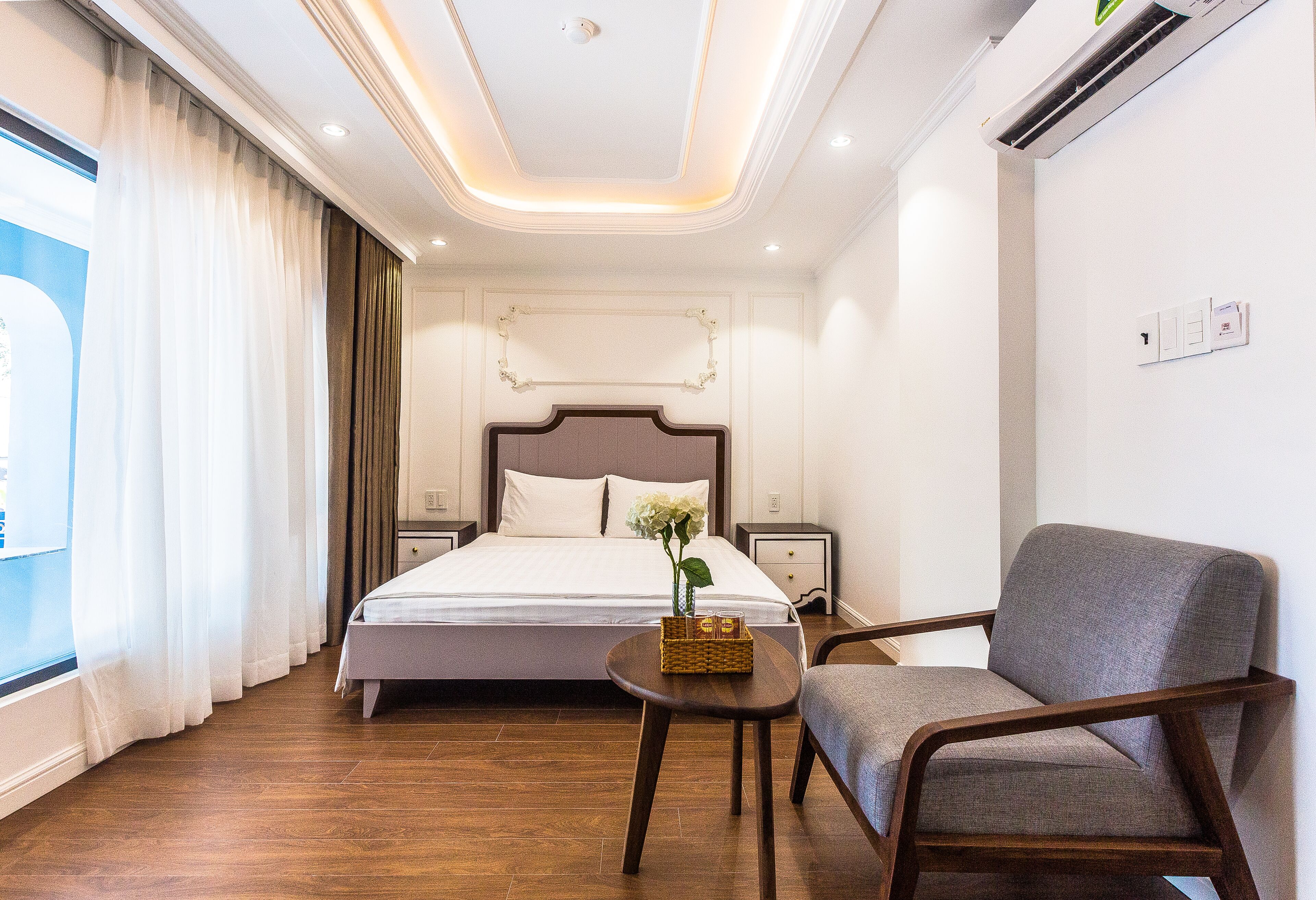 Quarto de hotel com cama grande, uma cadeira, mesa pequena com um vaso e janela com cortinas.