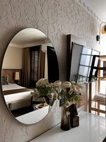Quarto de hotel com um espelho grande e redondo, uma cama, uma mesa de cabeceira com um vaso de flores e uma TV de tela plana fixada na parede.