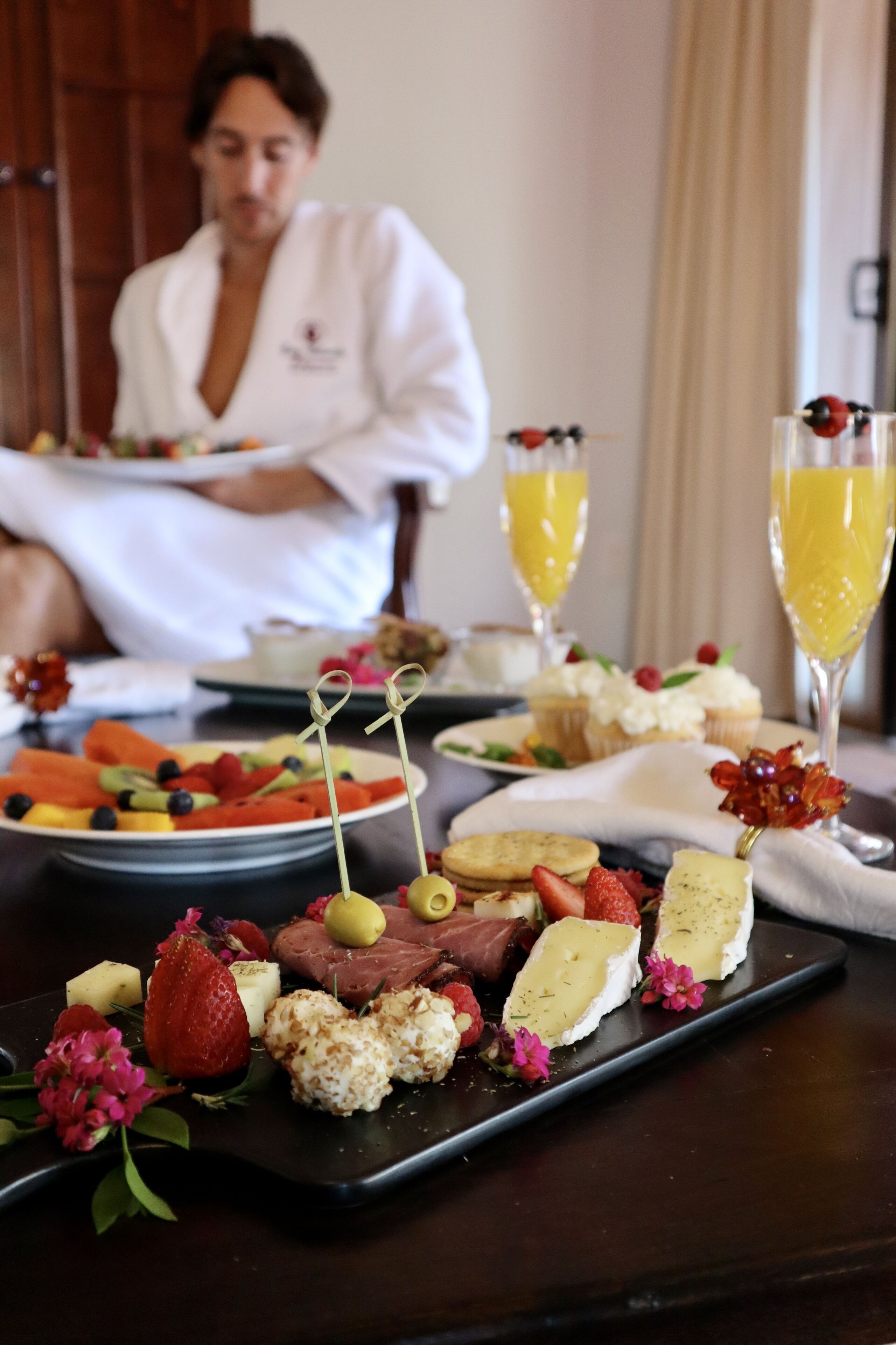 Mesa de café da manhã com diversas opções de alimentos, incluindo frutas, queijos e doces, além de dois copos de suco de laranja.