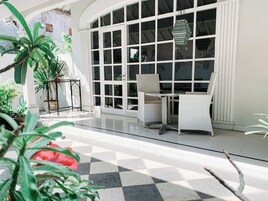 Um pátio banhado pelo sol, com piso xadrez, janelas com moldura branca, móveis de vime e plantas em vasos.