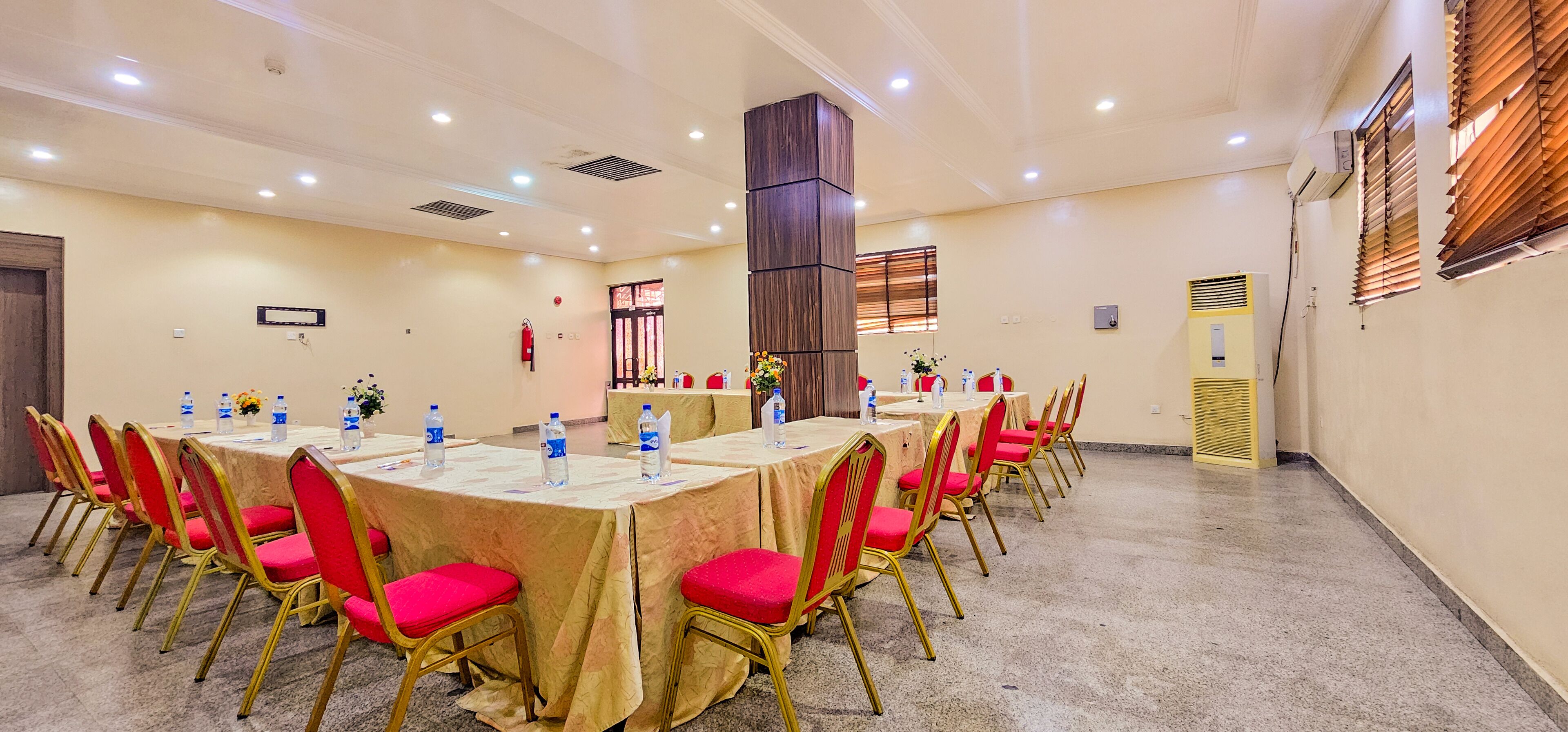 Sala de conferência com uma mesa comprida, cadeiras vermelhas e garrafas de água.
