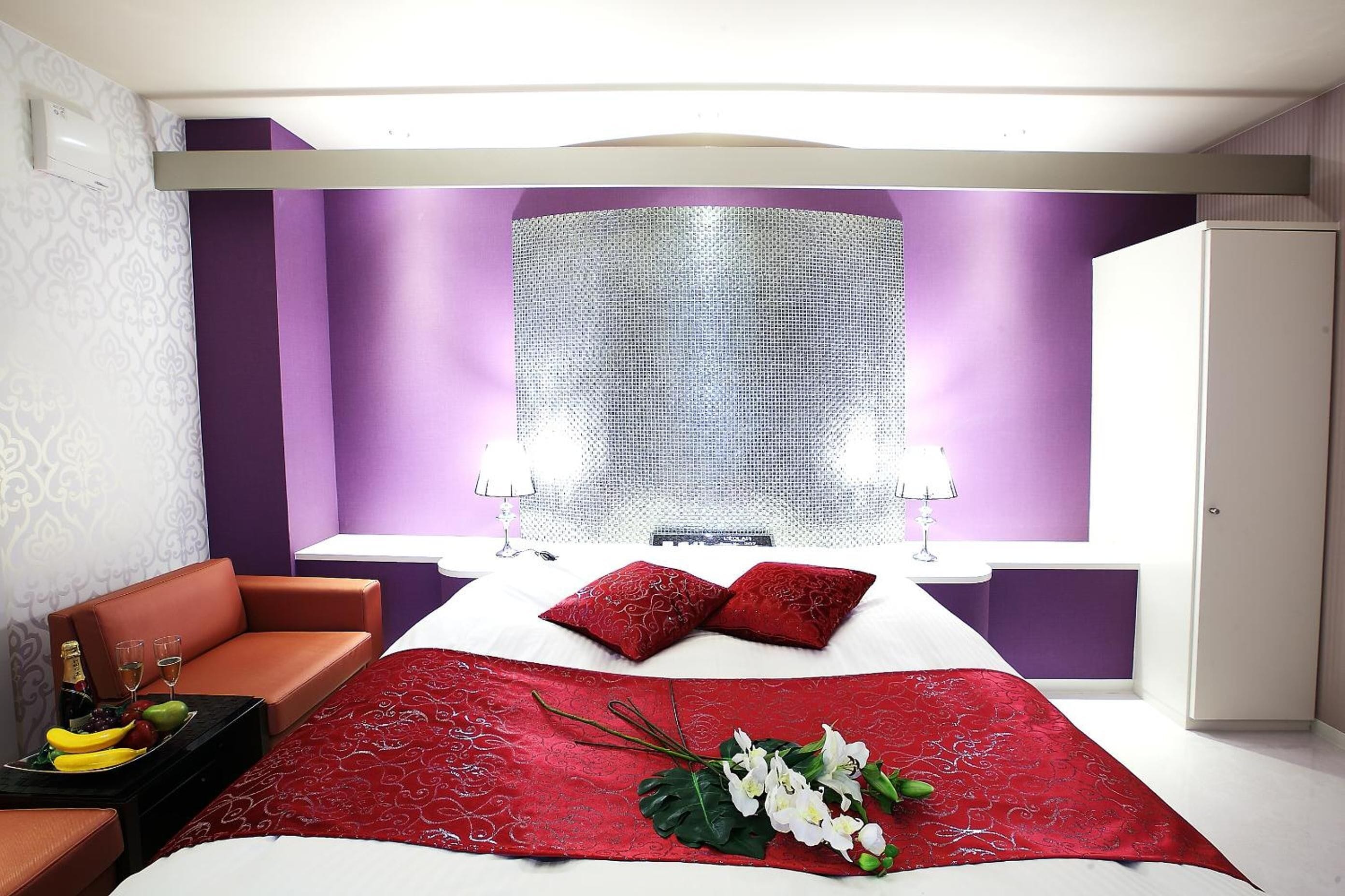 Quarto de hotel moderno com uma cama grande, uma manta vermelha, duas almofadas vermelhas, um arranjo de flores, uma pequena mesa com frutas e uma garrafa, e um guarda-roupa branco.