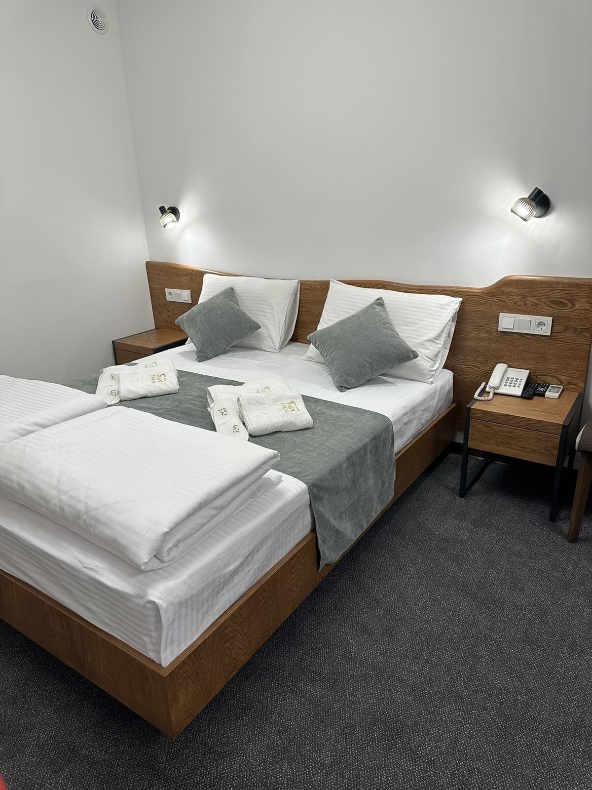 Quarto de hotel com cama de madeira, roupa de cama cinza, mesa de cabeceira com telefone e luminária fixada na parede.