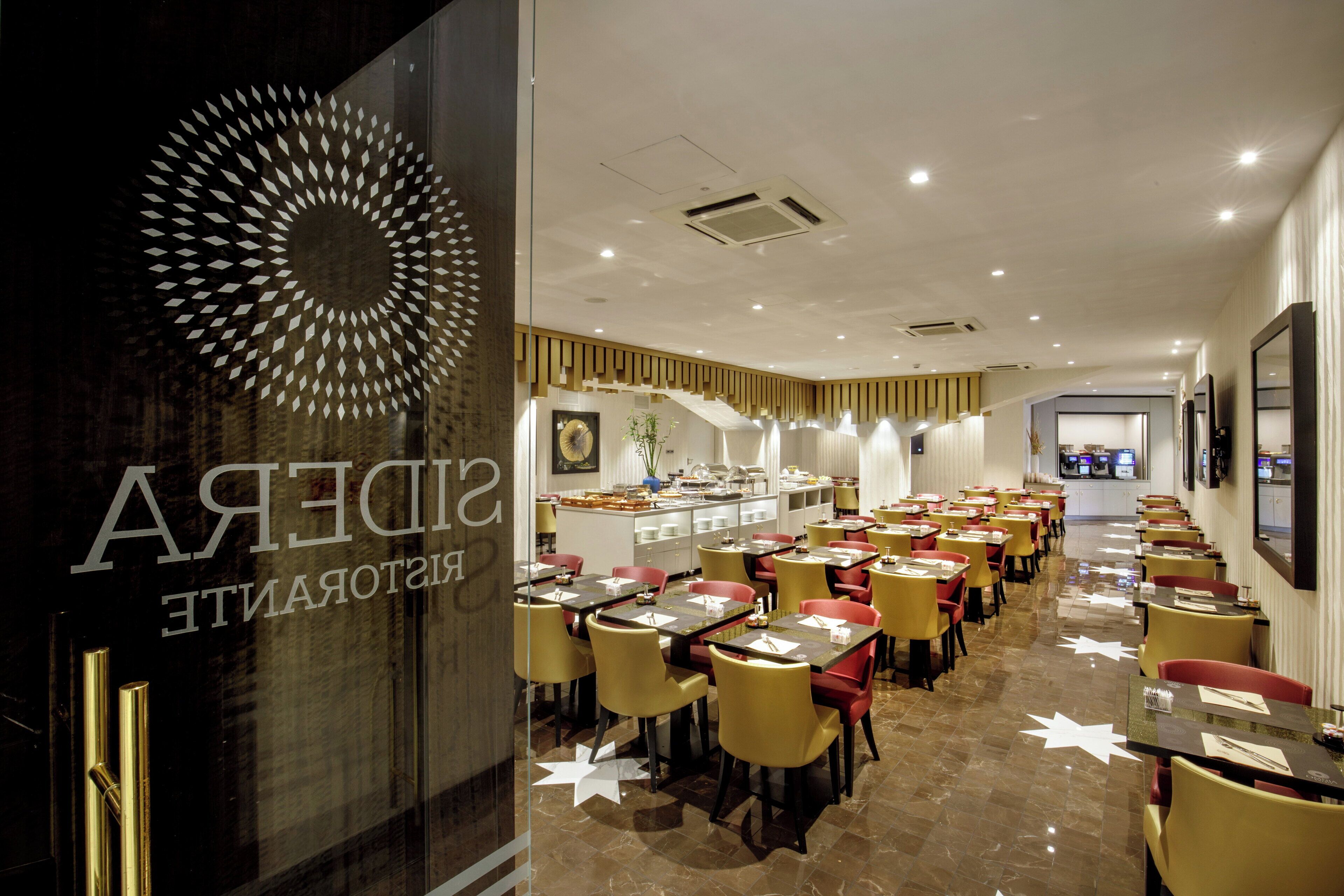 Um restaurante com interior moderno, apresentando uma área de jantar central com mesas e cadeiras, um balcão com vários pratos e uma escadaria que leva a um andar superior.