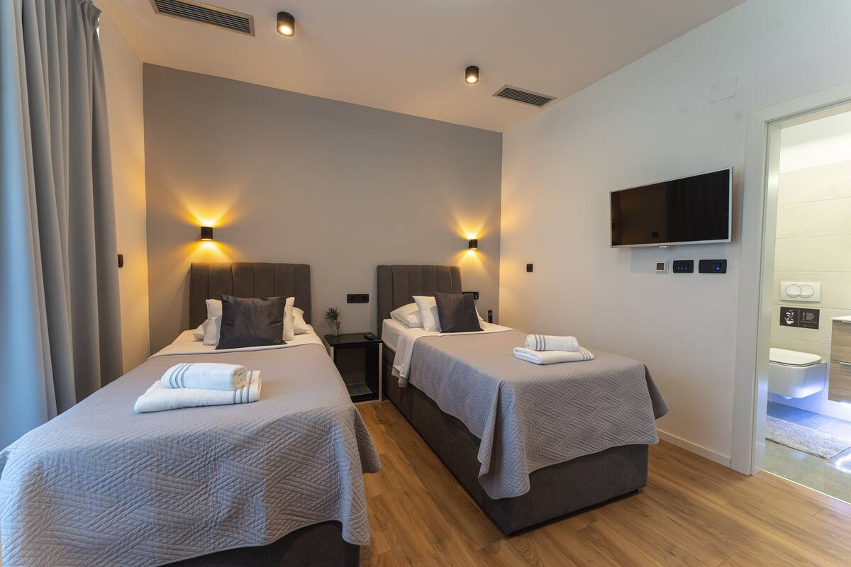 Quarto de hotel com duas camas, cada uma com cabeceira, mesas de cabeceira e uma TV de tela plana presa na parede.