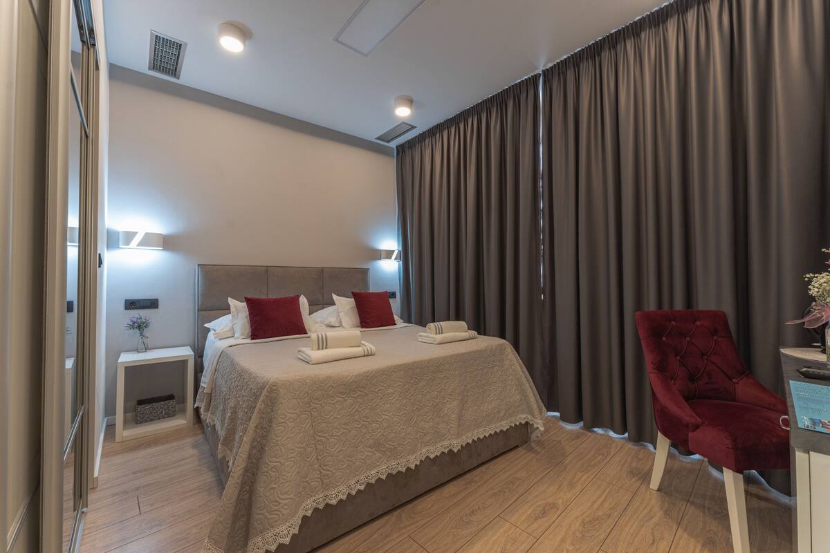 Quarto de hotel com uma cama grande, uma cadeira vermelha, uma pequena mesa de cabeceira com um vaso e um criado-mudo.