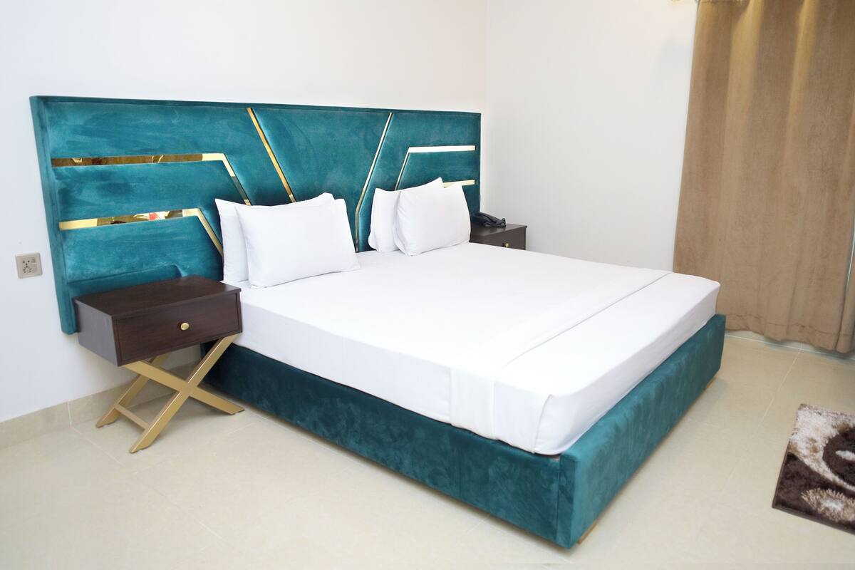 Quarto moderno com cabeceira estofada em tons de verde-água, mesa de cabeceira de madeira, colcha branca e um pequeno tapete no chão.