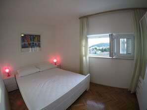 Um quarto com cama, mesa de cabeceira, janela com cortinas e um quadro na parede.