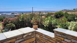 Um muro de pedra com uma planta em vaso, vegetação exuberante e vista para uma vila costeira com edifícios e o mar ao fundo.