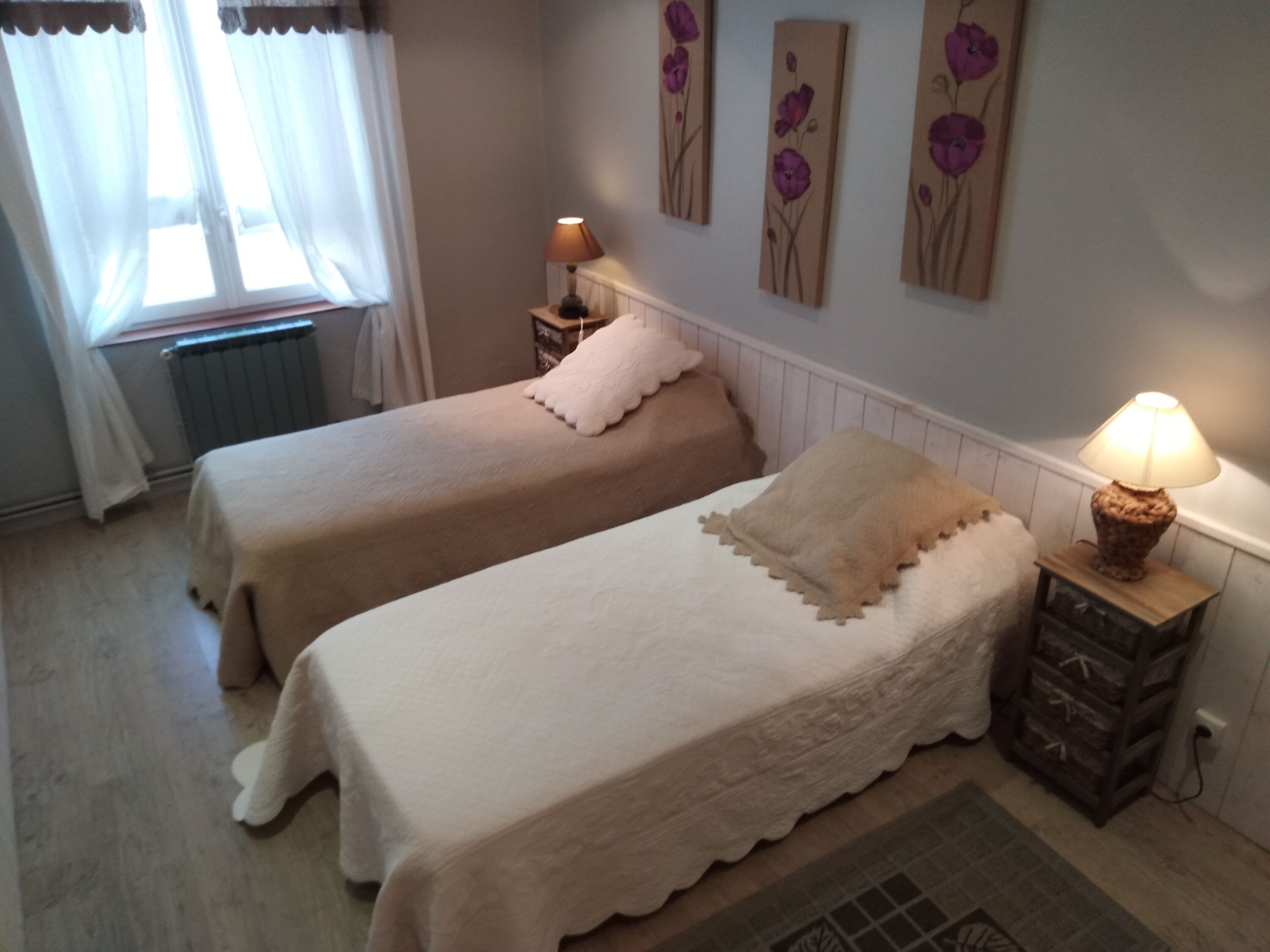 Quarto com duas camas de solteiro, mesa de cabeceira com abajur, radiador e janela com cortinas.