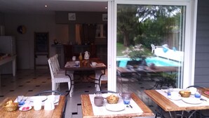 Uma área de refeições ao ar livre com mesas e cadeiras de madeira, piscina ao fundo e vista para o interior da casa.