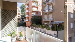 Uma varanda com vista para edifícios vizinhos, uma grade de metal e plantas em vasos.