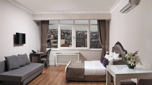 Quarto de hotel com sofá, cama, escrivaninha e vista para a paisagem urbana através de grandes janelas.