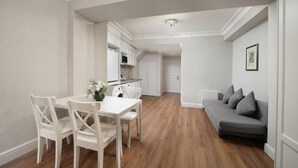 Uma cozinha moderna com armários brancos, uma sala de jantar com mesa e cadeiras brancas e um sofá cinza numa sala de estar com tons neutros.