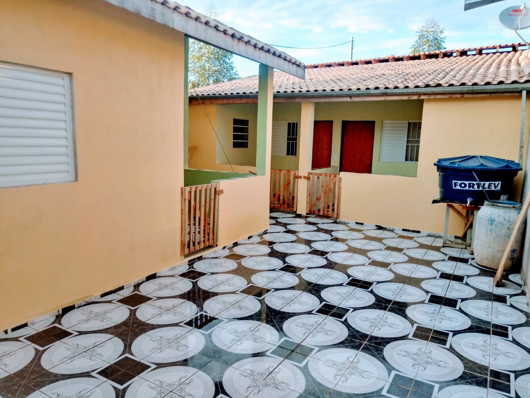 Um pátio com piso de azulejos, um barril azul identificado com 'FORTLEV' e uma antena parabólica no telhado.