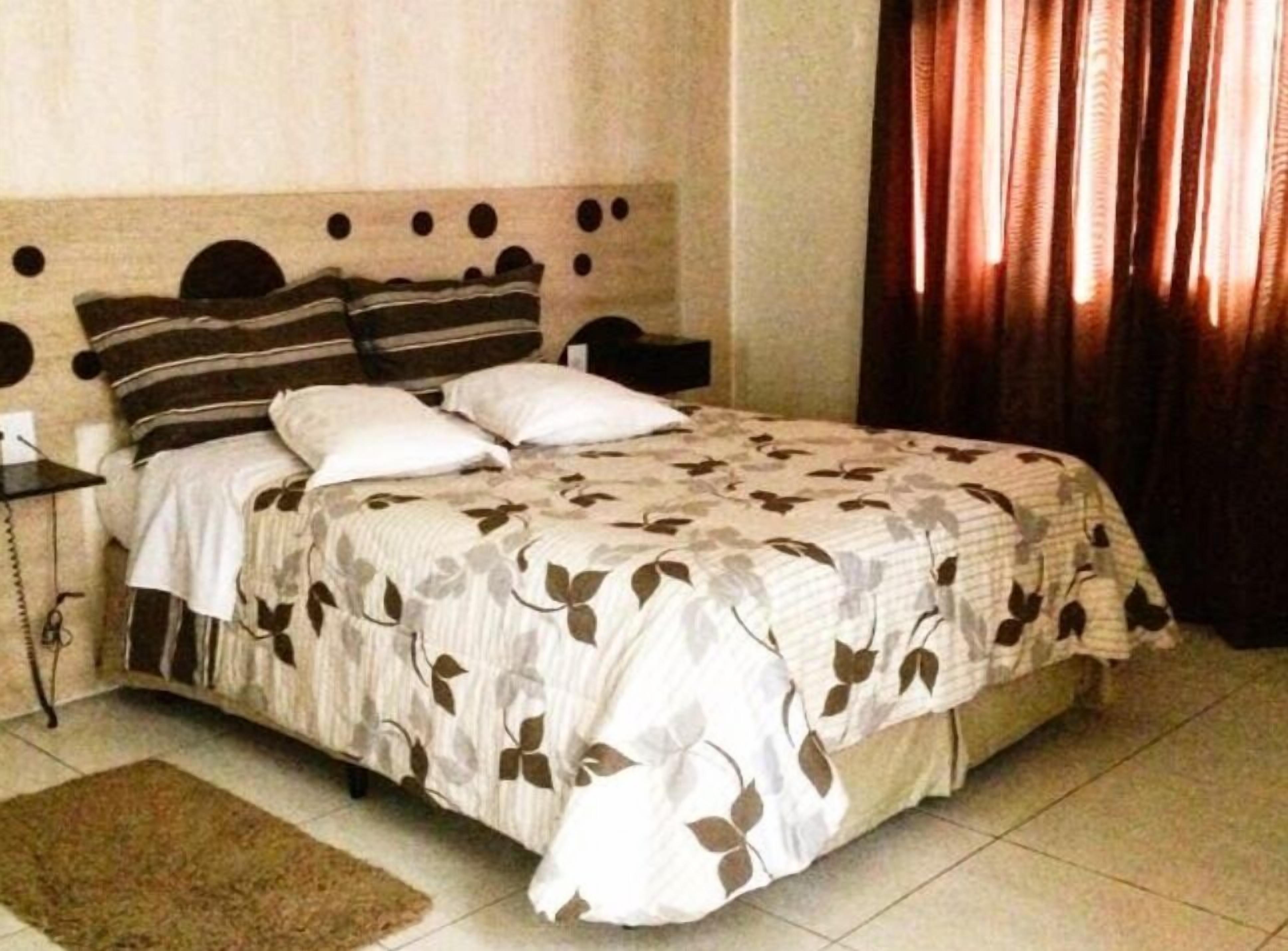 Uma cama bem arrumada com colcha e almofadas estampadas com flores, uma cabeceira de madeira, uma mesa de cabeceira com abajur e um tapete marrom no chão.
