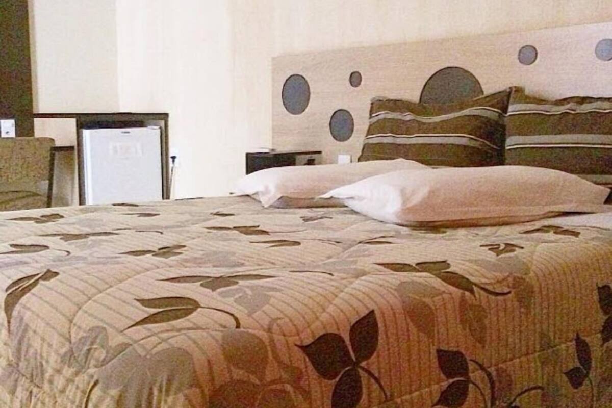 Quarto de hotel com uma cama, uma colcha estampada com padrão de folhas, uma mesa de cabeceira e um painel decorativo circular na parede.