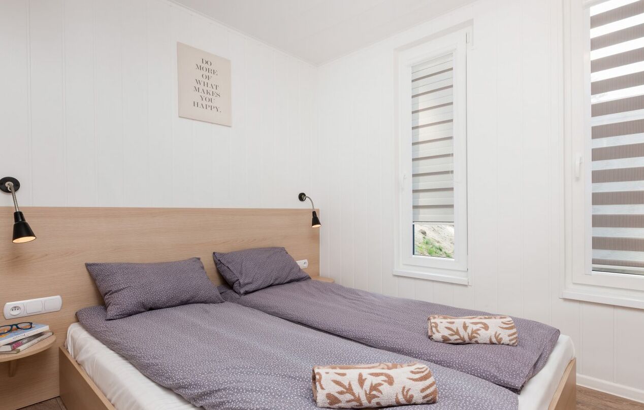 Uma cama de casal com cabeceira de madeira, duas luminárias de apoio e uma janela com persianas.
