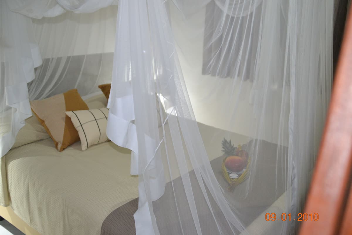 Quarto com cama coberta por um dossel branco, uma mesinha com uma fruteira e uma janela com cortinas leves.