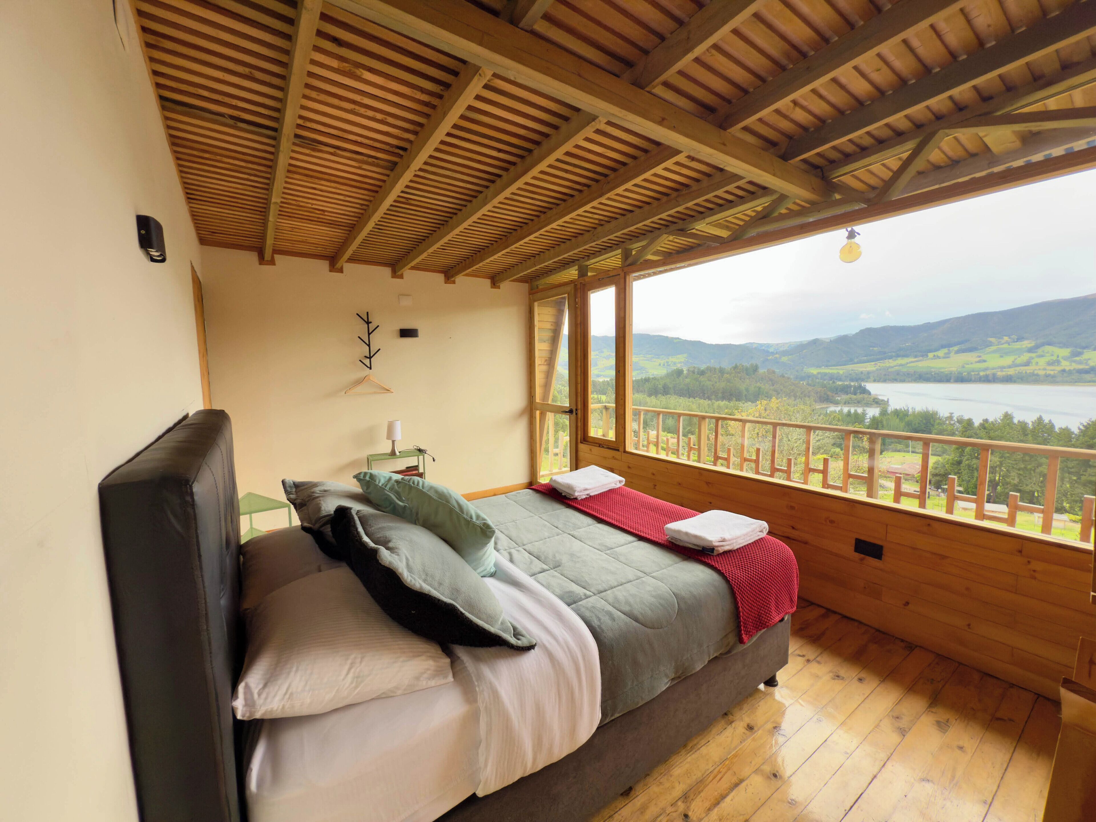 Um quarto com cama, almofadas, mesa de cabeceira, abajur e vista para um lago e montanhas.