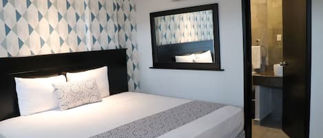 Quarto de hotel com cama, cabeceira, parede estampada, ar condicionado e banheiro visível através de uma porta aberta.