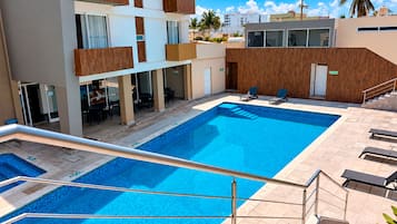 Uma piscina com deck de madeira, cadeiras de descanso e uma escadaria que leva a um edifício com varandas.