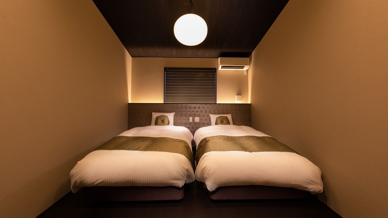 Quarto de hotel estreito com duas camas, uma lâmpada no teto e um ar condicionado fixado na parede.