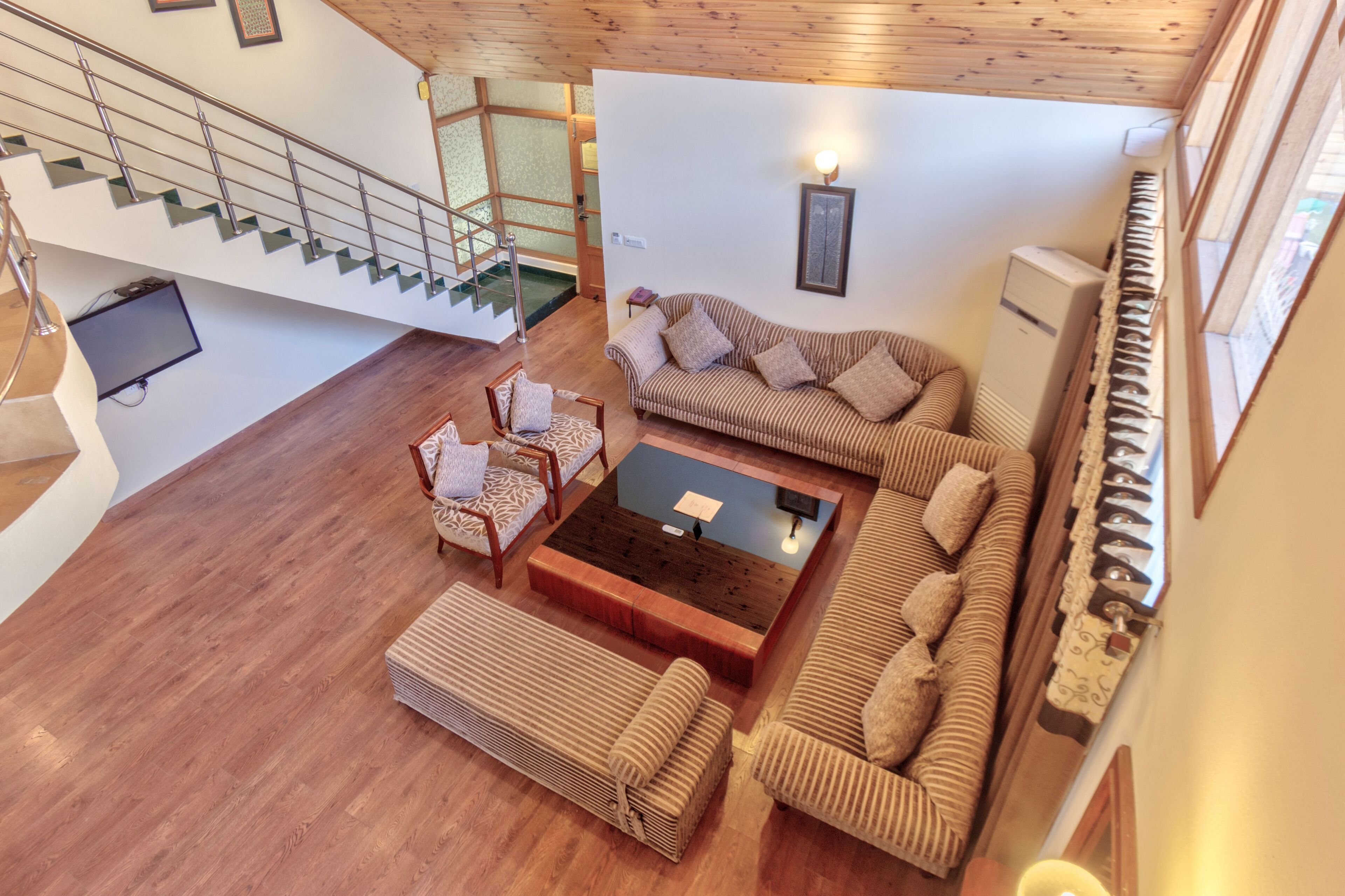Uma sala de estar ampla com escada de madeira, sofá, mesa de centro e televisão na parede.