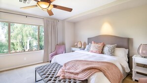 Um quarto com uma cama grande, um banquinho, um ventilador de teto, uma mesa de cabeceira e uma janela com cortinas.