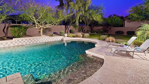 Piscina com borda xadrez, cercada por um jardim paisagístico com arbustos e árvores.