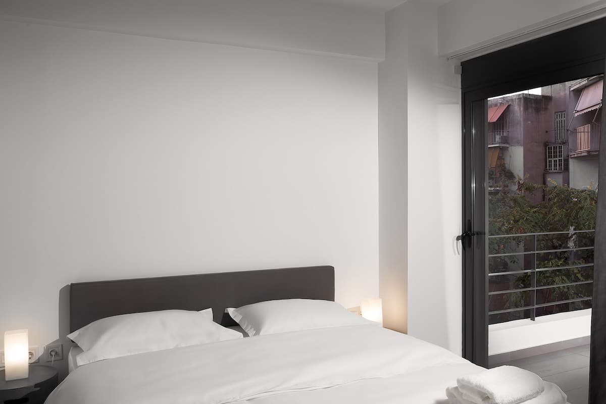 Quarto de hotel moderno com uma cama, uma mesinha de cabeceira e vista para o exterior através de uma porta de vidro.