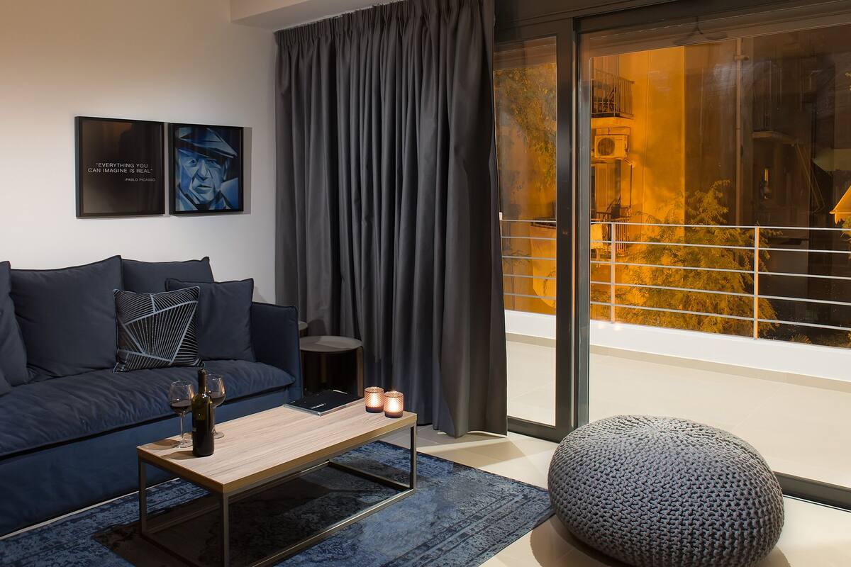 Uma sala de estar moderna com um sofá azul escuro, uma mesa de centro de madeira e um banquinho redondo. Há duas frases emolduradas na parede e uma janela grande com cortinas.