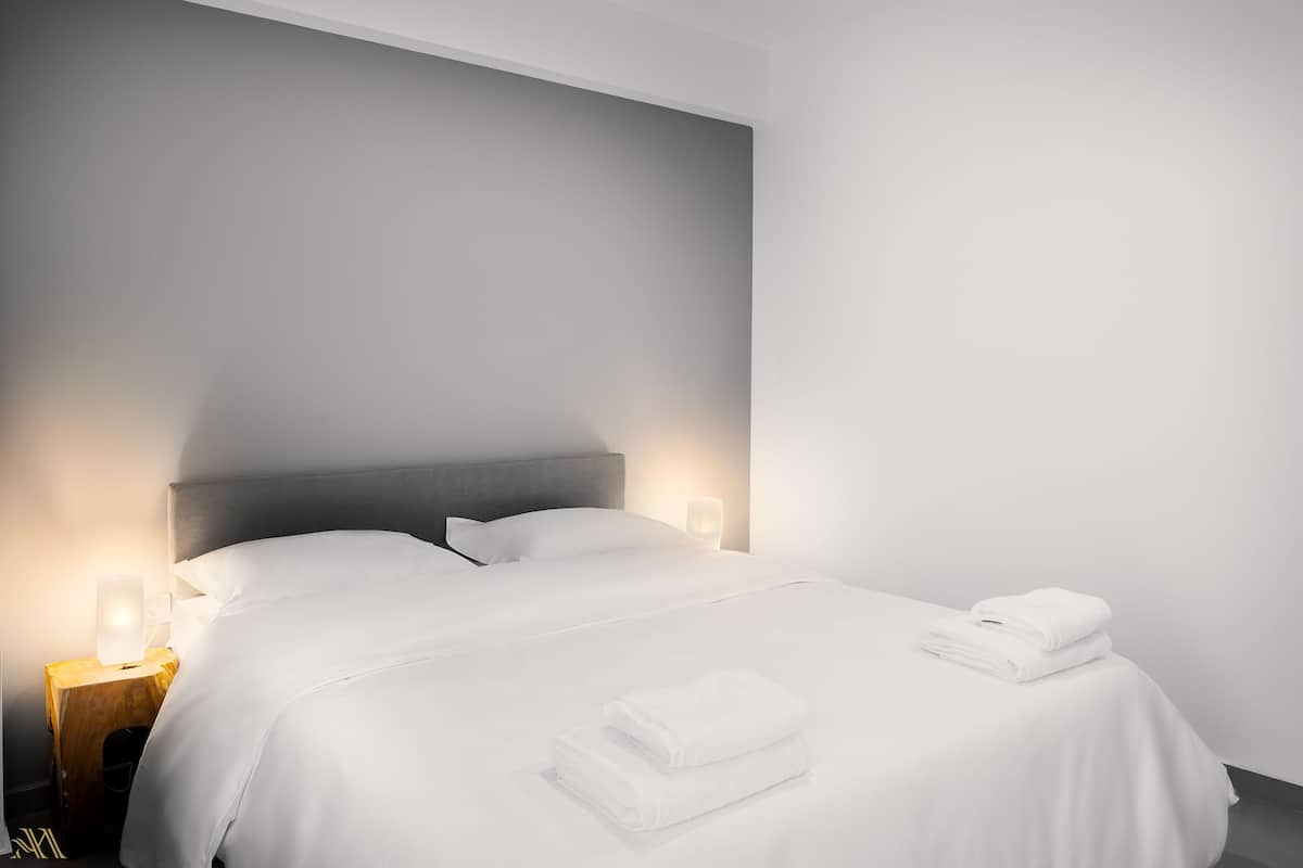 Cama bem arrumada com lençóis e toalhas brancas, mesa de cabeceira de madeira com abajur e cabeceira moderna.