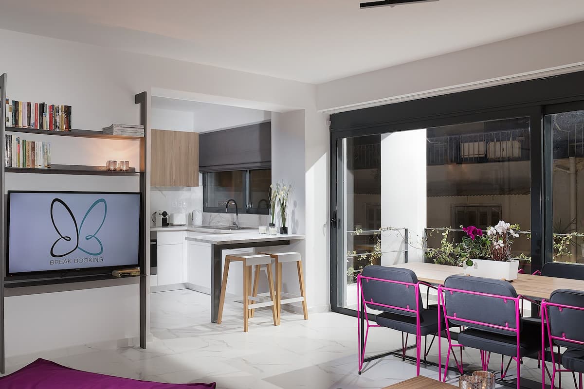 Uma sala de estar moderna com televisão de tela plana, uma estante, um espaço de refeições com cadeiras cor-de-rosa e uma cozinha ao fundo.