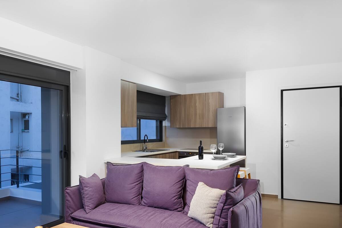 Uma sala de estar moderna com um sofá roxo, uma área de cozinha com geladeira e uma porta de vidro deslizante que dá para uma varanda.