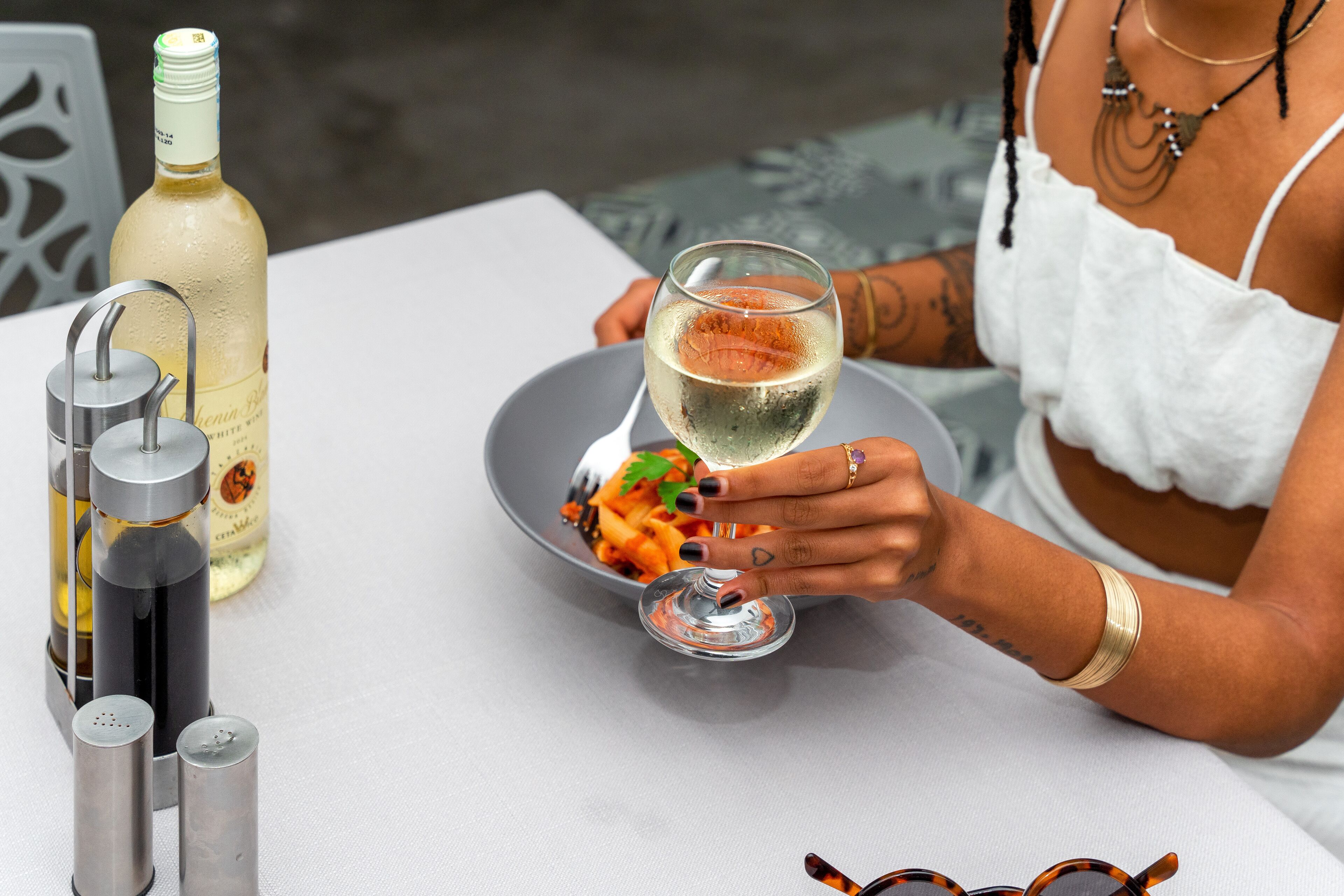 Uma pessoa segurando uma taça de vinho branco, sentada à mesa com um prato de comida, uma garrafa de vinho branco e saleiro e pimenteiro.