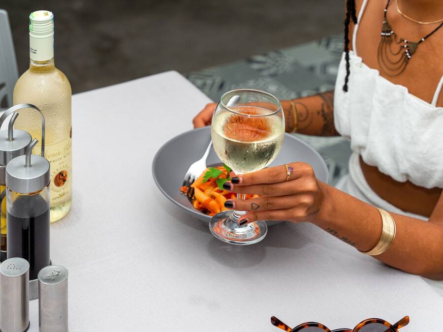 Uma pessoa segurando uma taça de vinho branco, sentada à mesa com um prato de comida, uma garrafa de vinho branco e saleiro e pimenteiro.