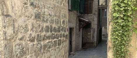 Um beco estreito com calçada de pedra, com uma parede de pedra rústica de um lado e um edifício com uma persiana verde do outro.