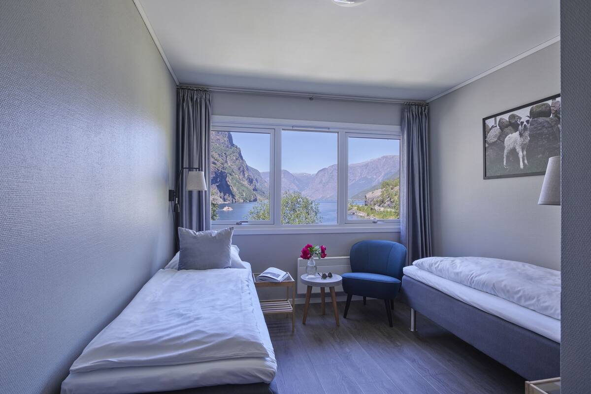 Quarto de hotel com duas camas de solteiro, uma mesinha com abajur, uma cadeira e vista para uma paisagem montanhosa.