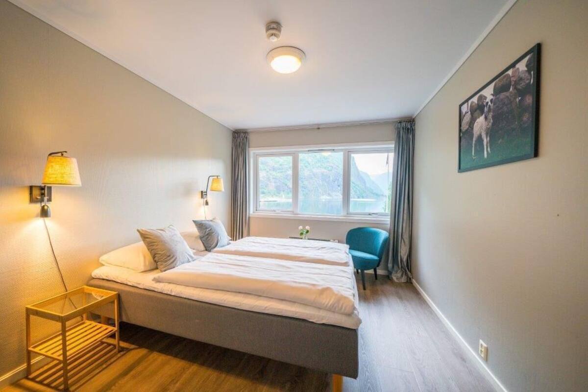 Quarto de hotel com cama, abajures, uma cadeira, janela com cortinas e um quadro na parede.